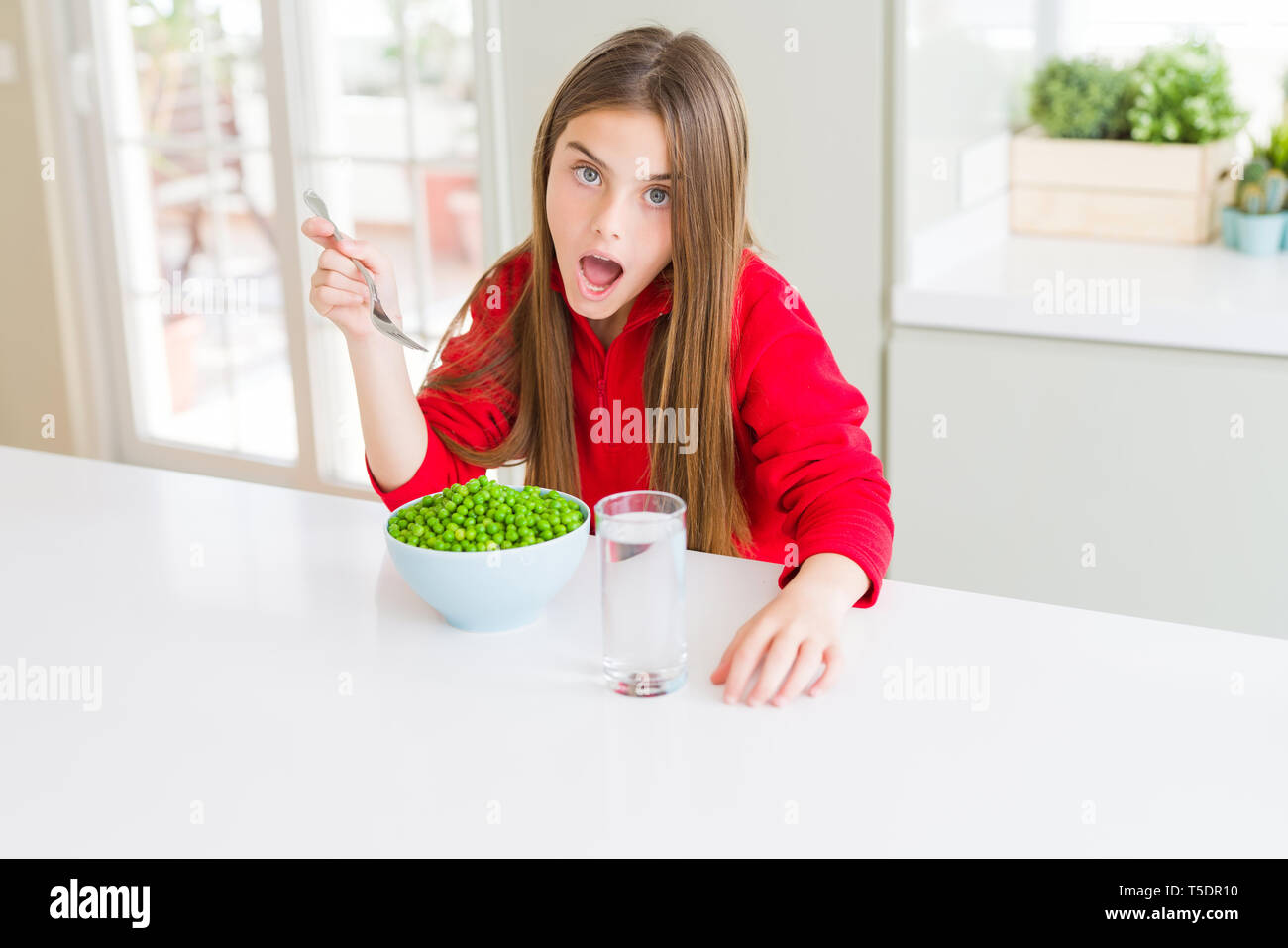 Beautiful young girl eating healthy green peas scared in shock with a ...