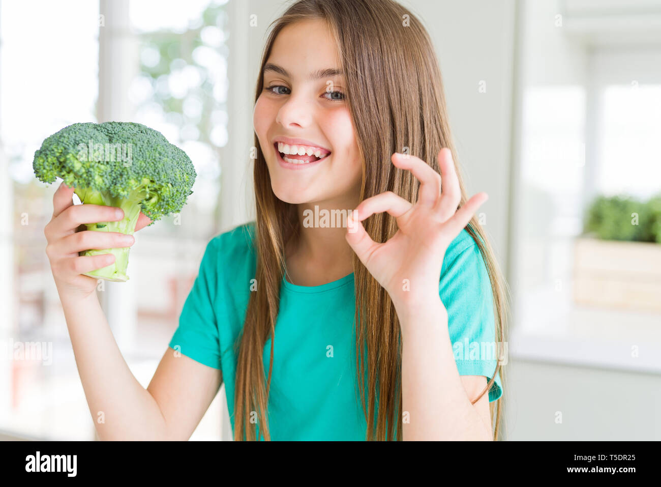 Beautiful young girl eating fresh broccoli doing ok sign with fingers ...