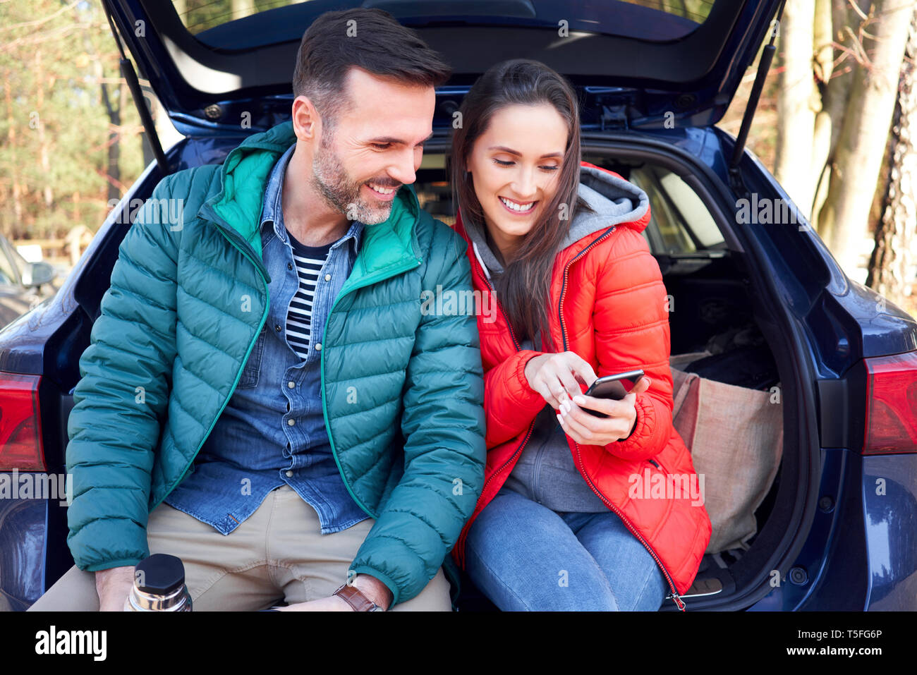 Couple using smartphone while sitting in car trunk during road trip ...