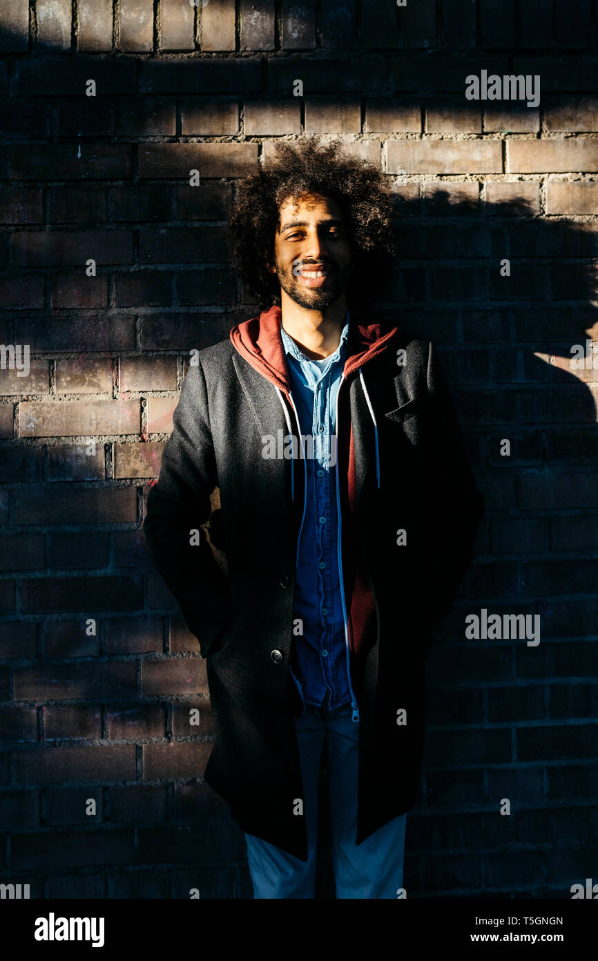 Portrait of a smiling young man standing at a brick wall in shadow ...
