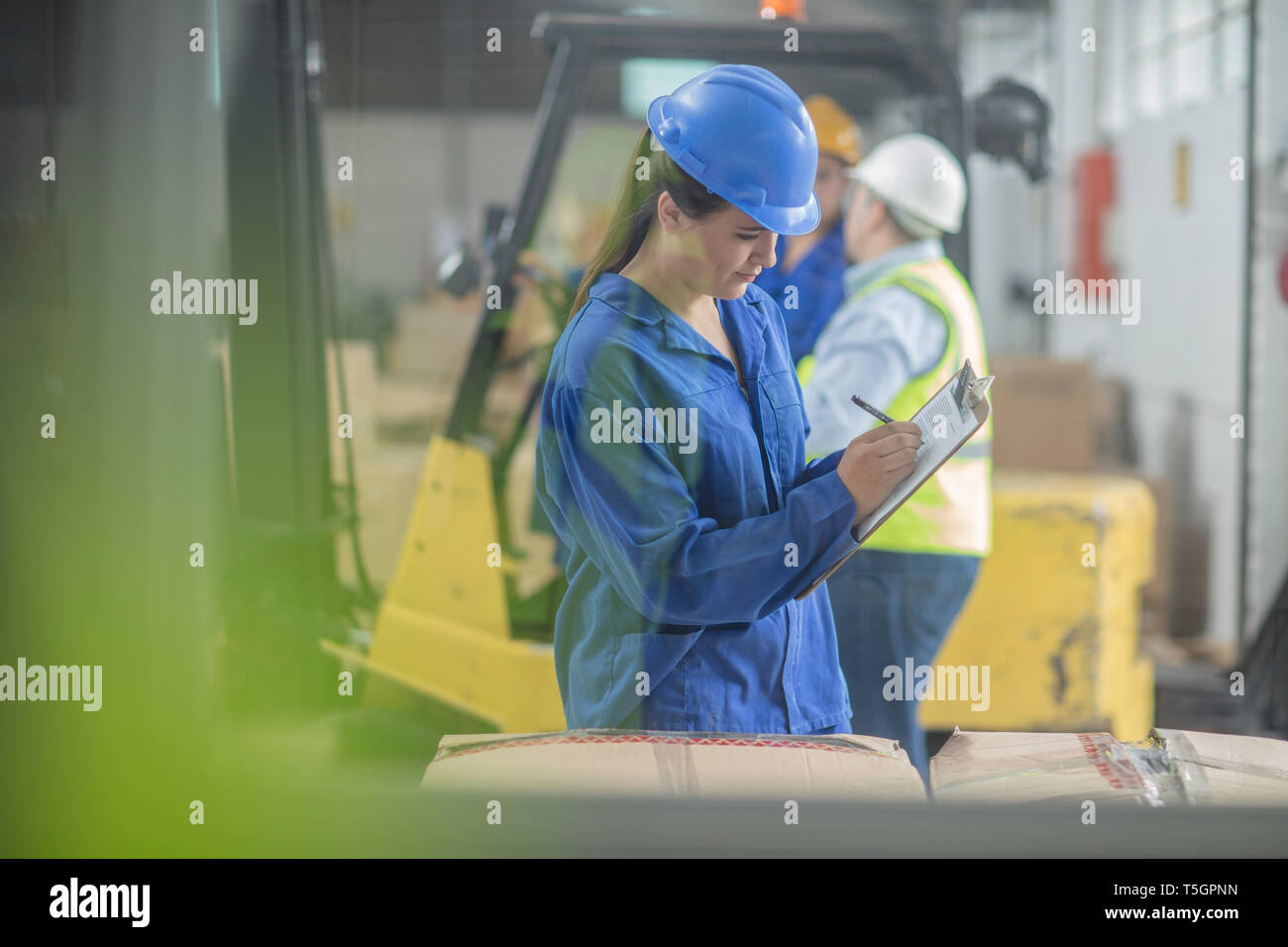 Woman wearing hard hat writing on clipboard in factory Stock Photo - Alamy