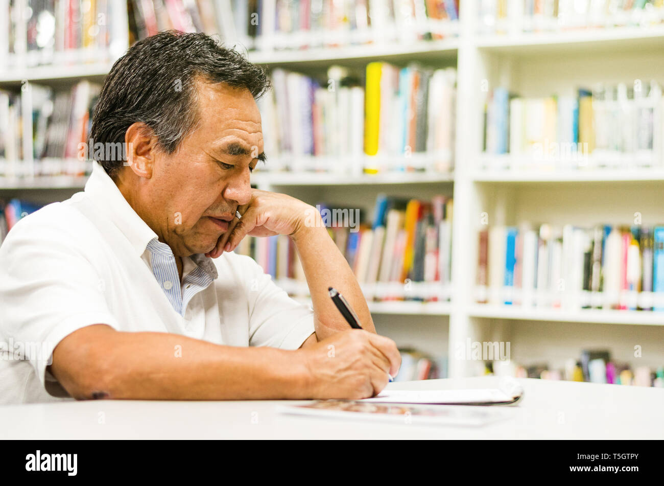 Serious senior man sitting on a library bench, writing in his book ...