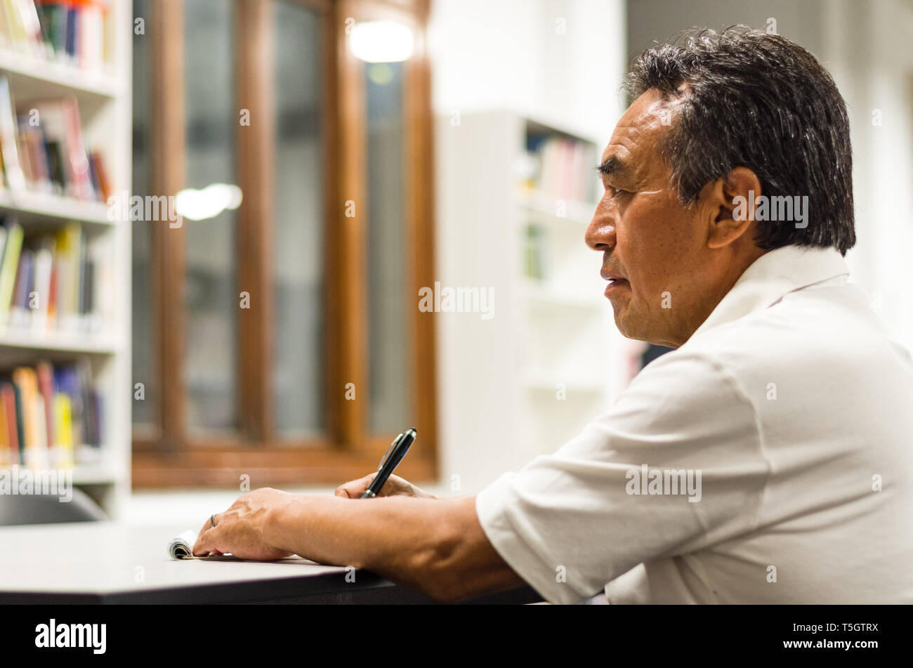 Serious senior man sitting on a library bench, writing in his book ...