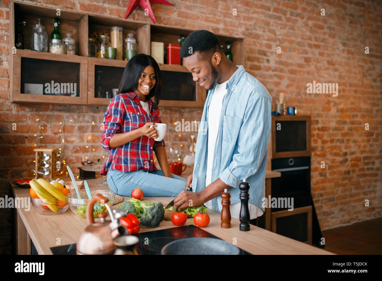Black man cooking breakfast on the kitchen, wife drinks coffee. African couple preparing ...