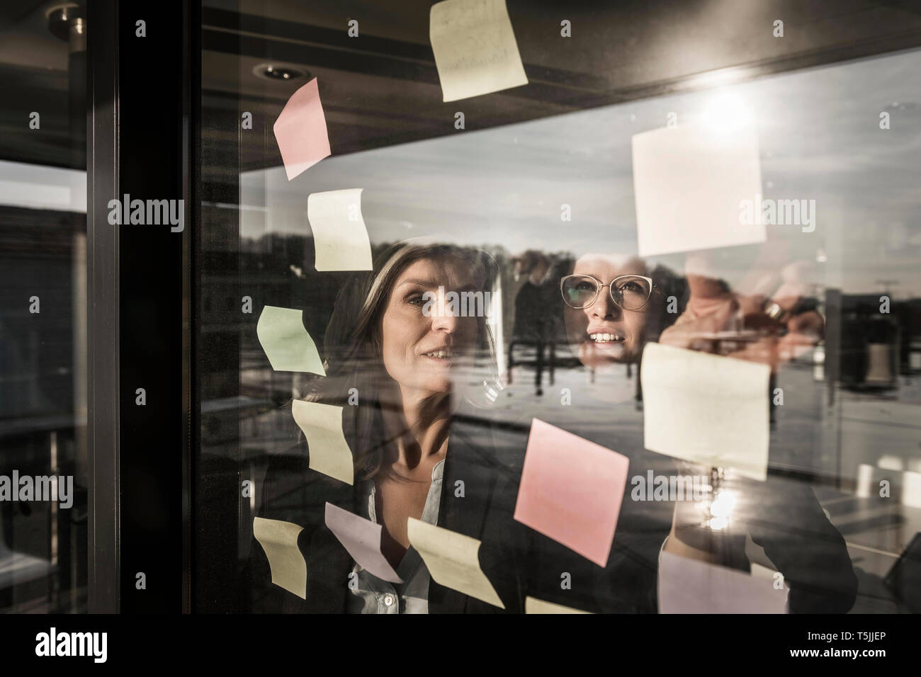 Two businesswomen brainstorming, putting sticky notes on window pane ...