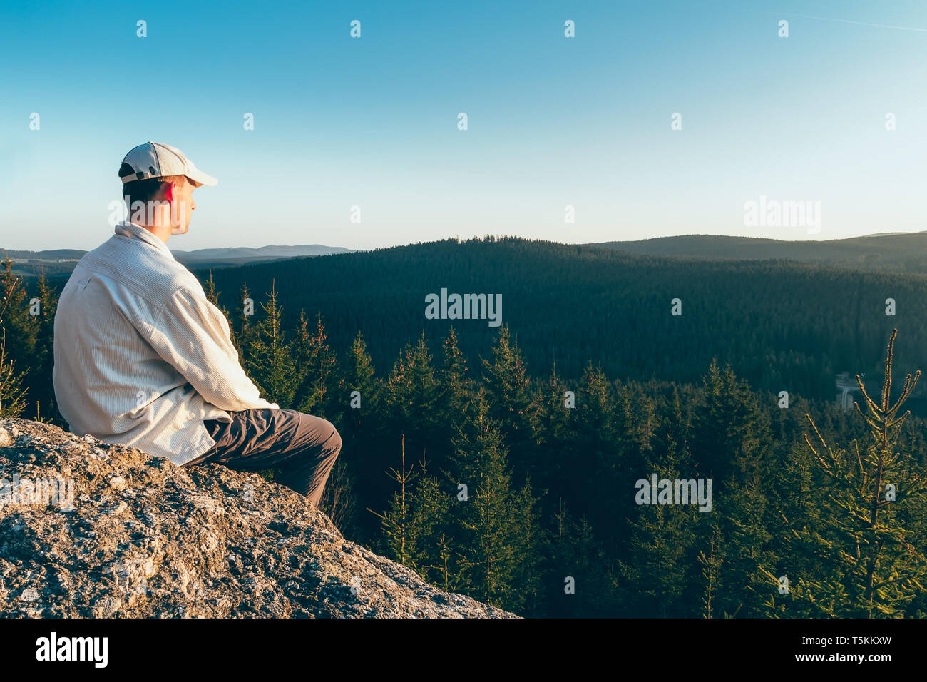 Young man hiker in white cap and shirt sit on rock from back looking to ...