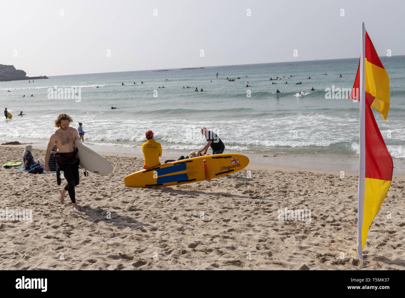 RNLI Lifeguard sat on surfboard at a busy Sennen Cove beach, Cornwall ...