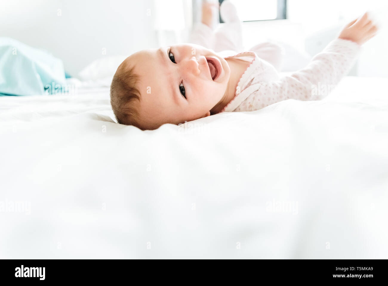 Happy and smiling adorable 6 month old baby girl lying on a bed, lifestyle isolated on natural ...