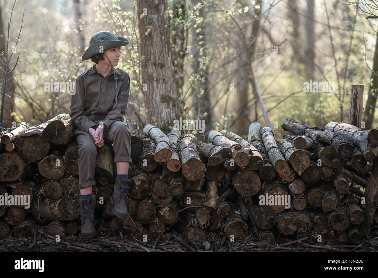 A boy in a coveralls and a hat sitting on a pile of sawn logs in the ...