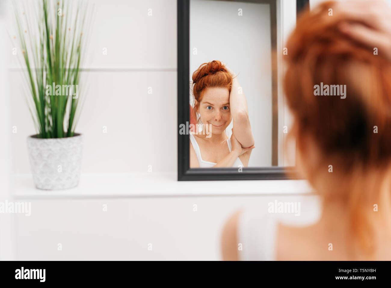 Cute young woman looking at herself in a mirror in a clean white ...