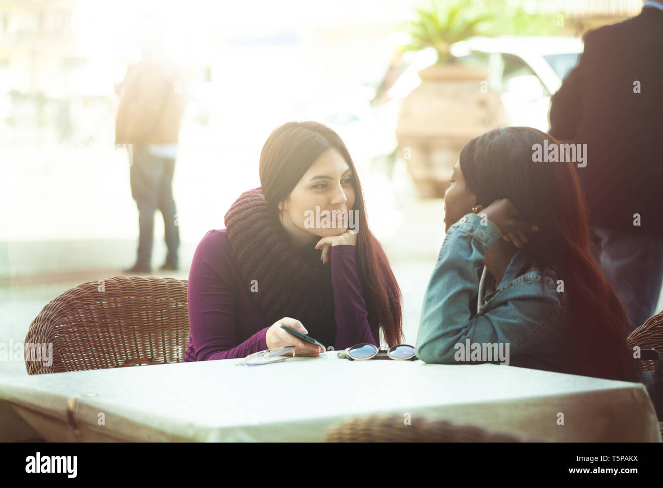Two young beautiful girls sit at a table talking and using the mobile ...