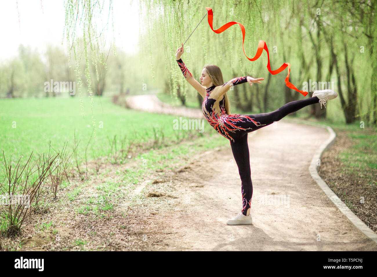 Rhythmic gymnastics. Young gymnast girl with red ribbon outdoor in park ...