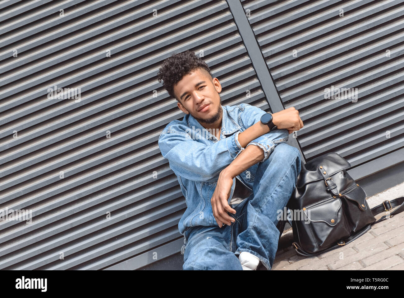 Young mulatto man sitting with leather backpack on city street leaning ...