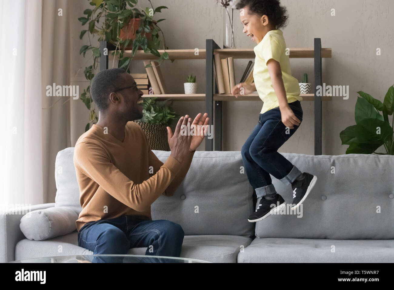 African father clapping hands looking at son jumping on sofa Stock ...