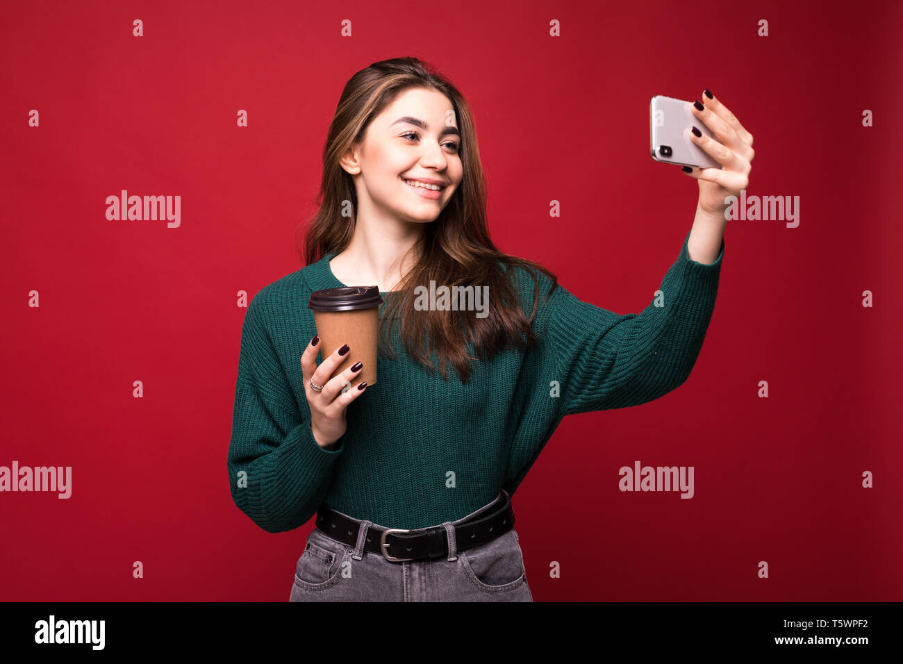Cool classy woman taking selfie on her cell phone with coffee cup isolated over red background ...