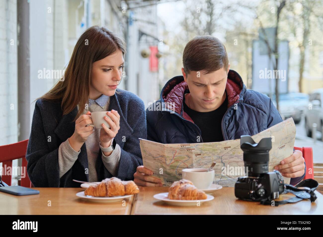 Young tourists man and woman reading map of city in outdoor cafe ...