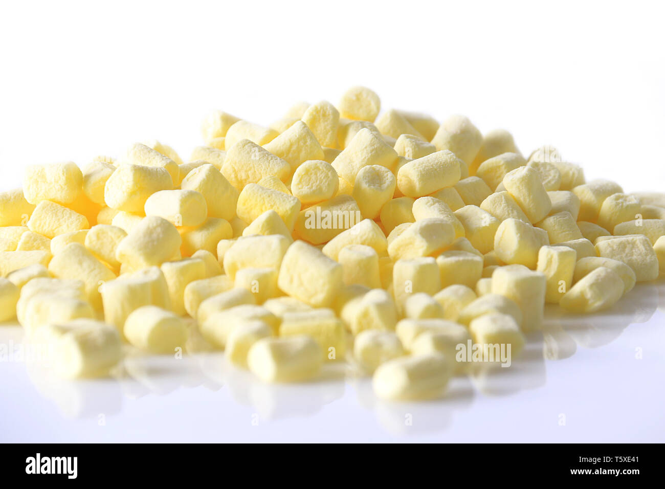 Pastel yellow mini marshmallows scattered on a white reflective surface ...