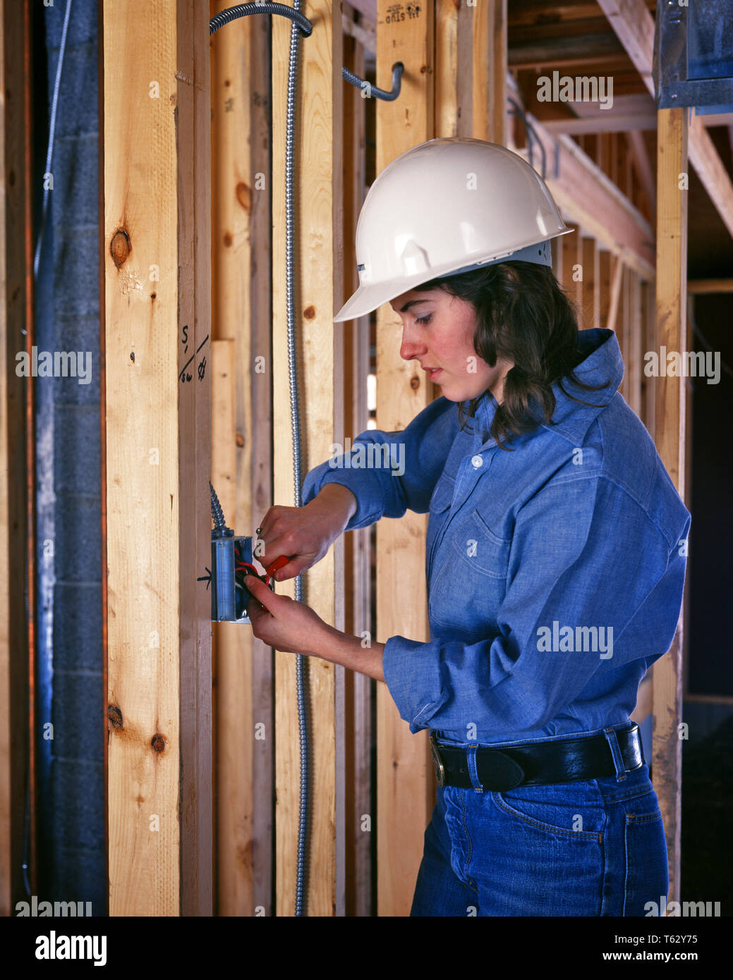 1970s YOUNG WOMAN ELECTRICIAN WEARING WHITE HARD HAT WORKING INSTALLING ...