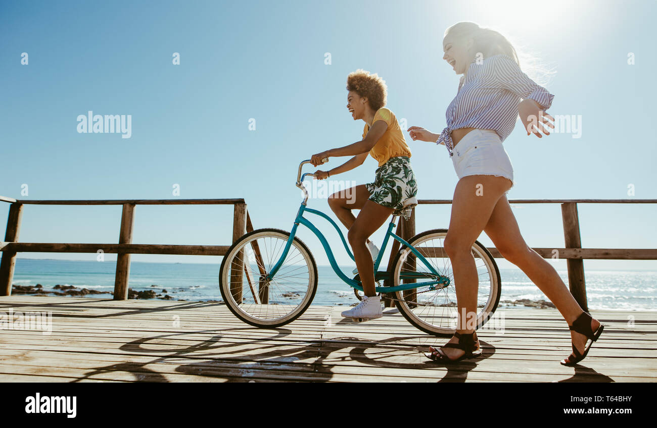Young woman riding a bicycle with her friend running by on boardwalk ...