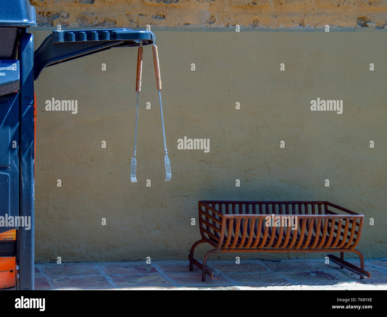 BBQ tongs hanging from a BBQ grill near to a rusted iron wood rack ...