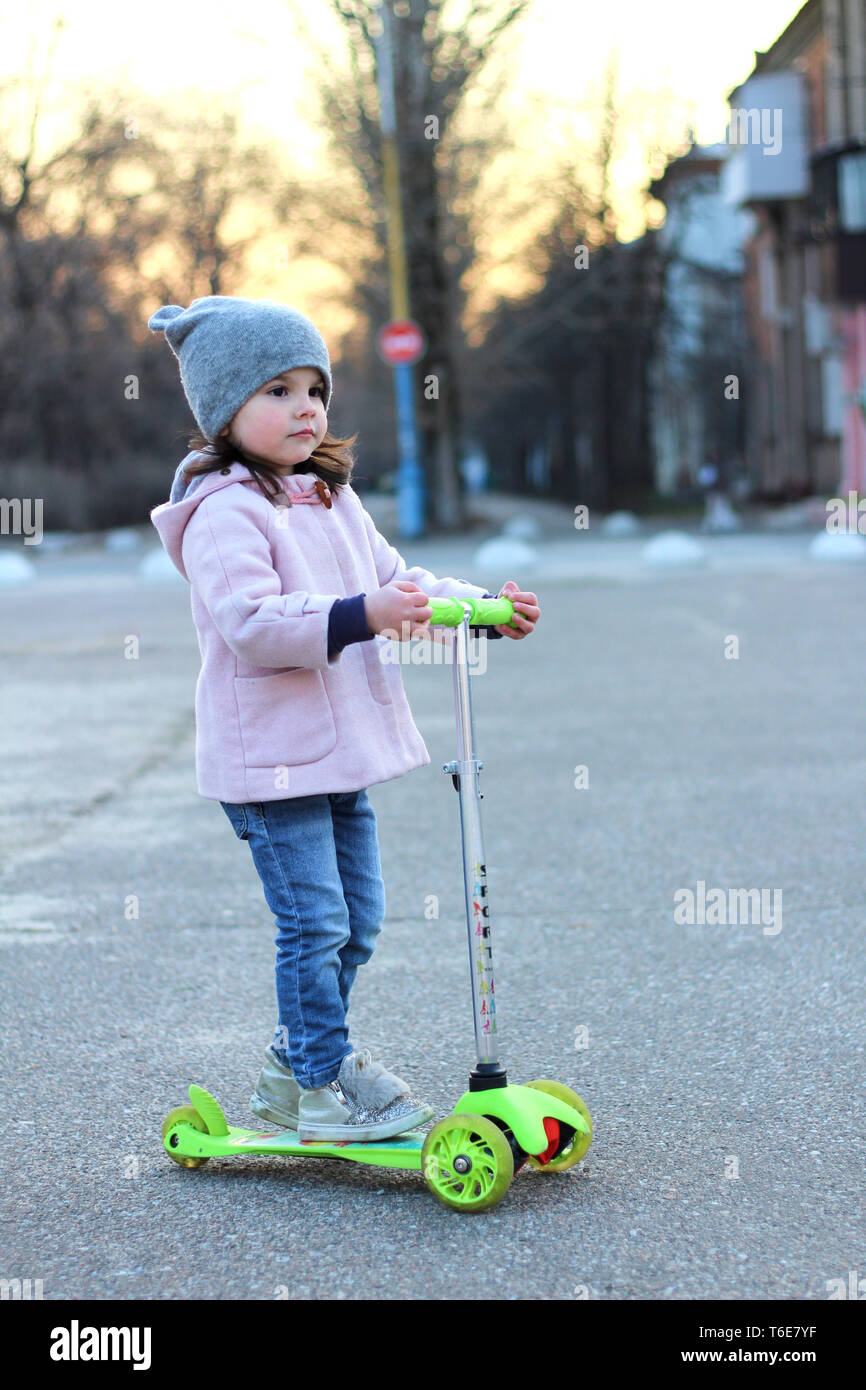 Cute girl in a hat, coat and jeans riding a three-wheeled scooter ...