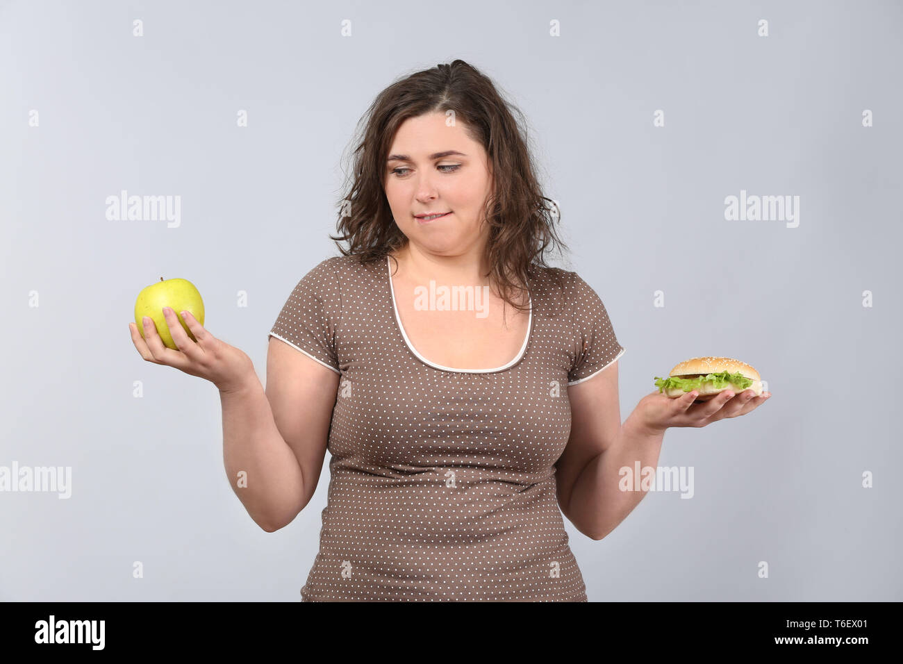 Young stout woman choosing between burger and apple on grey background ...