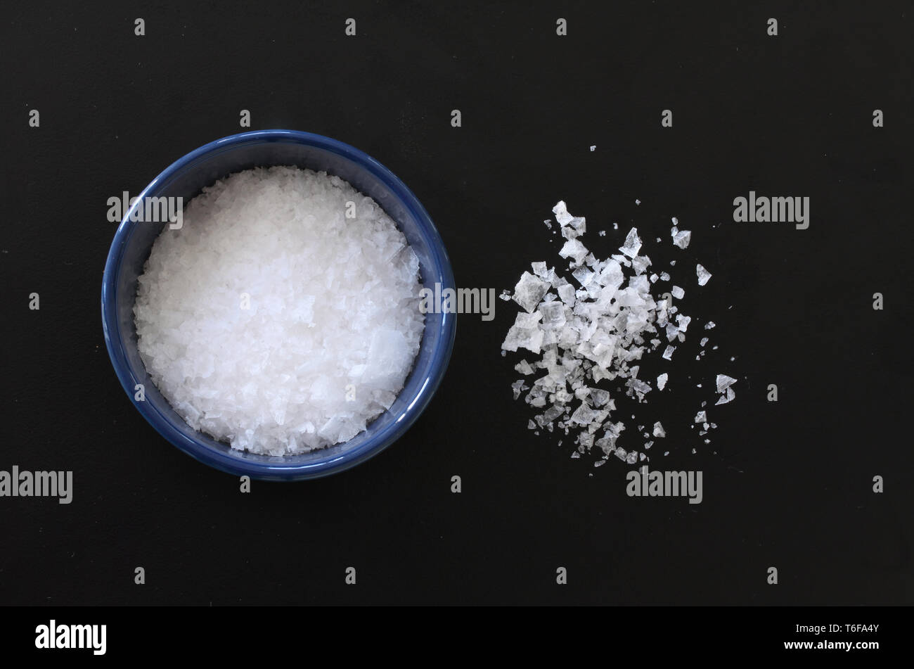 Flake salt lying in a blue bowl and scattered around on a black ...