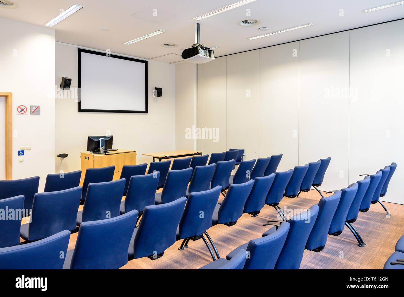 Empty presentation room with rows of blue seats, white projection screen, video projector on the ...