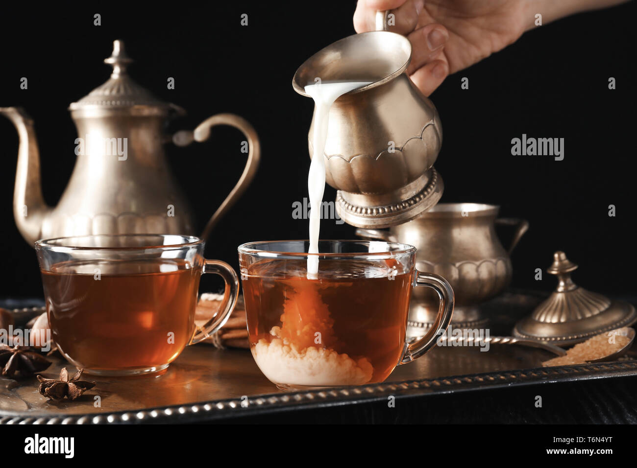 Pouring of milk into glass cup with tea on tray against dark background ...