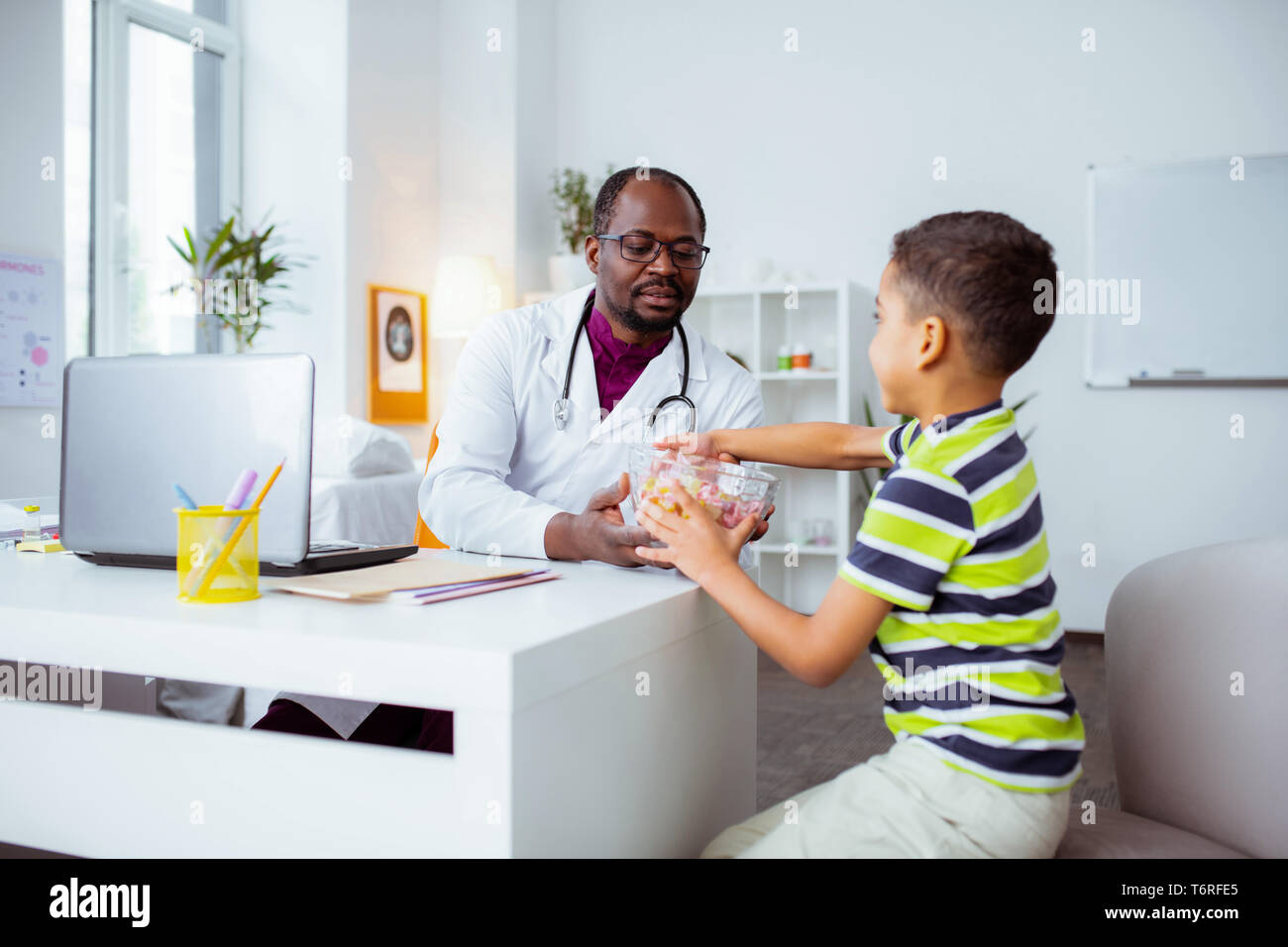 Dark-skinned doctor giving bowl with sweet candies to his lovely son ...