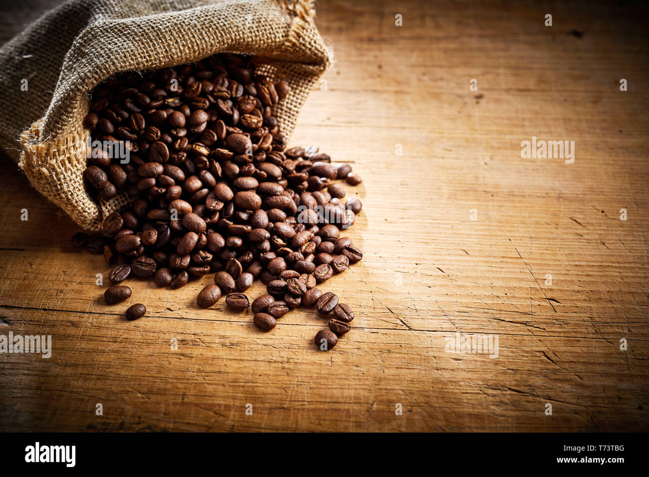Fresh roasted coffee beans spilling from a hessian sack onto a rustic wood background with copy ...