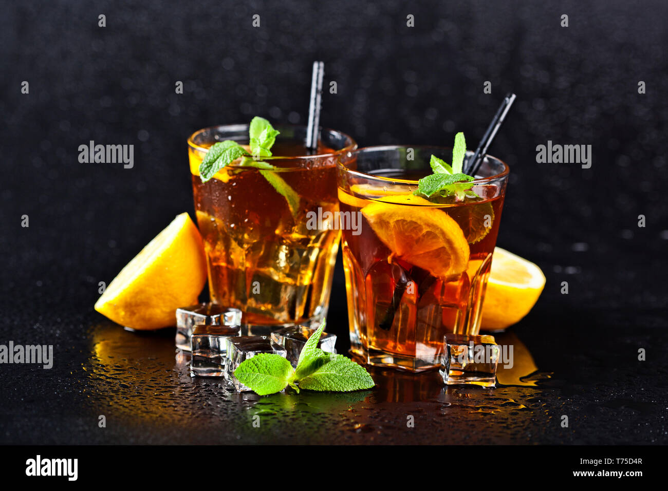 Two glasses with cold traditional iced tea with lemon, mint leaves and ice cubes on wet black ...