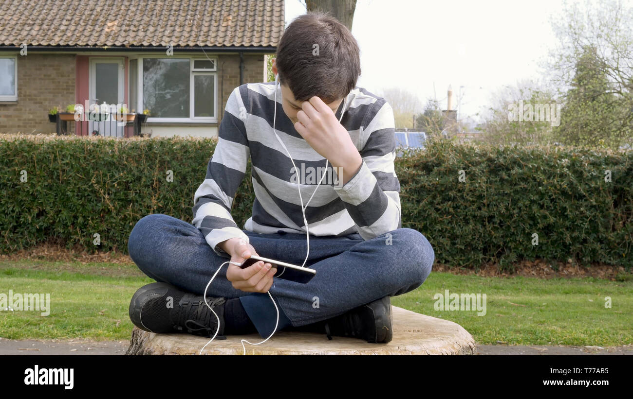 teen boy with smart phone listening or talking while sitting on stump ...