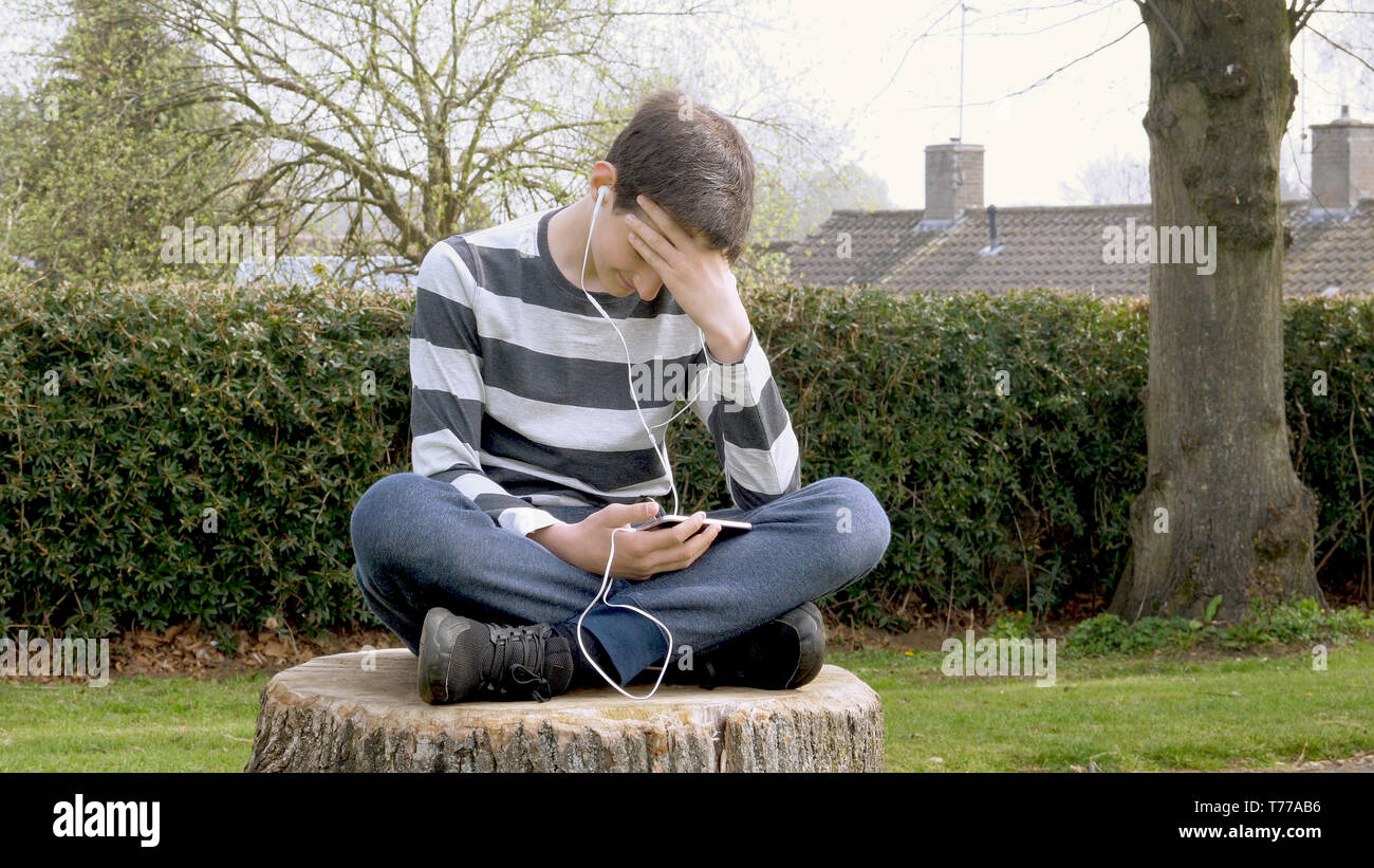 teen boy with smart phone listening or talking while sitting on stump ...