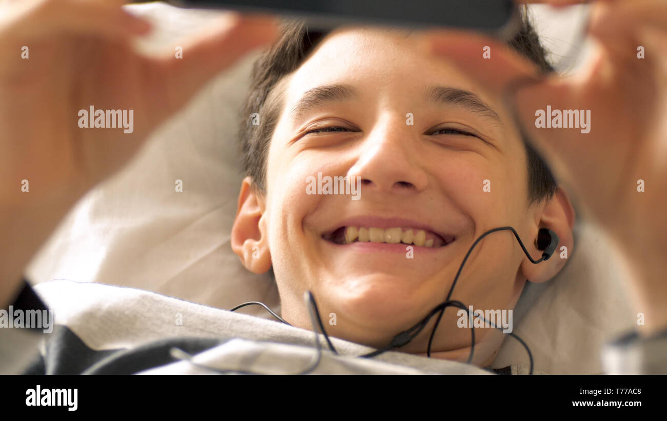 smiling teen boy with smart phone listening or talking on sofa indoor ...