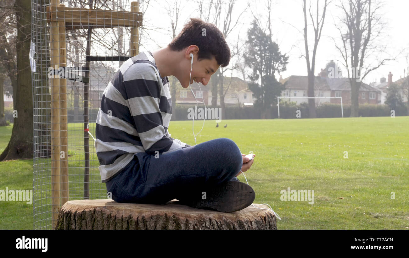 teen boy with smart phone listening or talking while sitting on stump ...