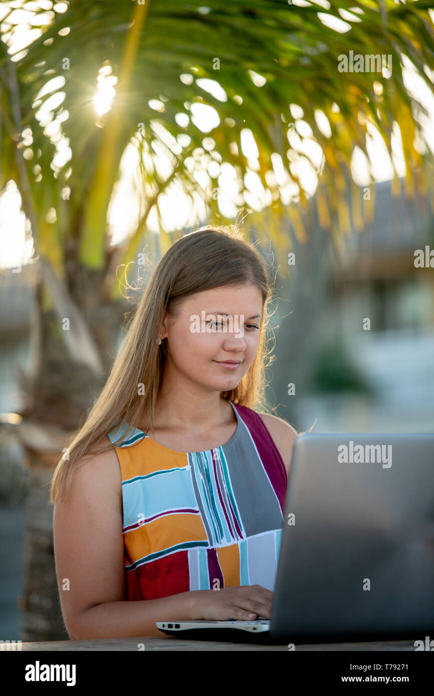 young woman wearing summer dress works with laptop computer under palm ...