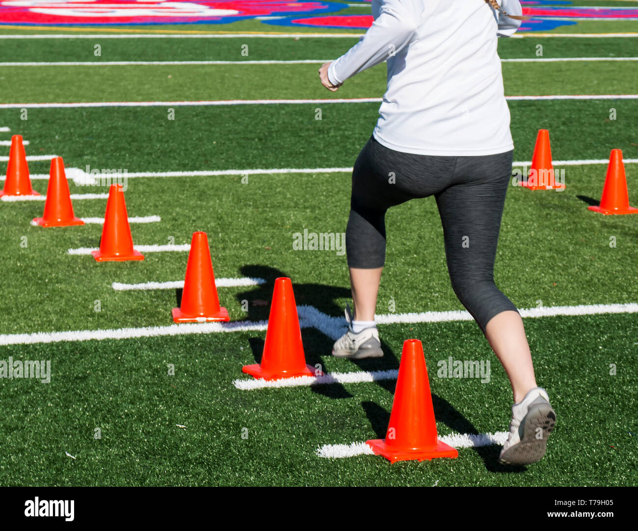 A female runner performing a speed drill over orange cones on a green turf field on a sunny ...