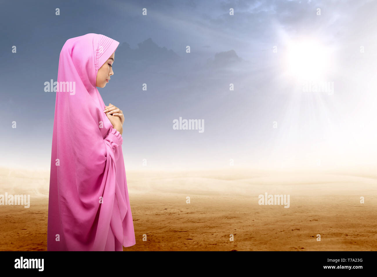 Asian muslim woman in veil praying on desert with sun rays and dark sky ...