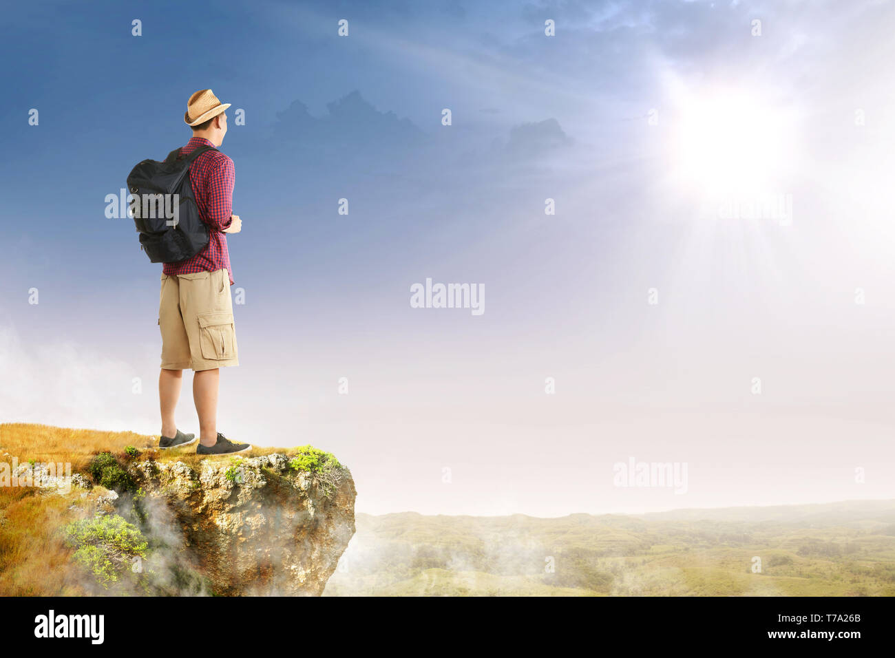 Rear view of asian traveler man with hat and backpack standing on the edge of cliff looking at ...