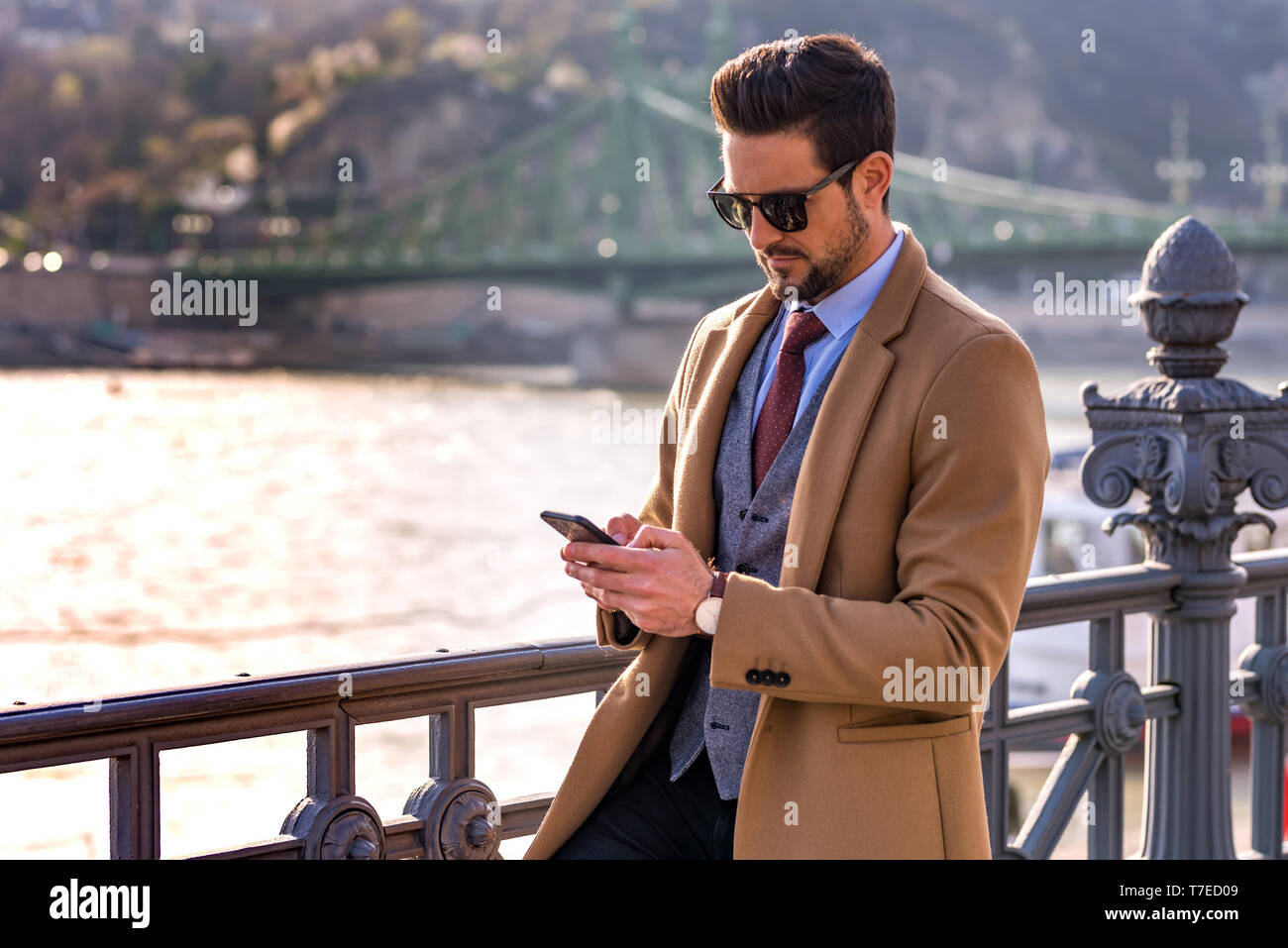 A handsome elegant man standing next to a river and using his ...