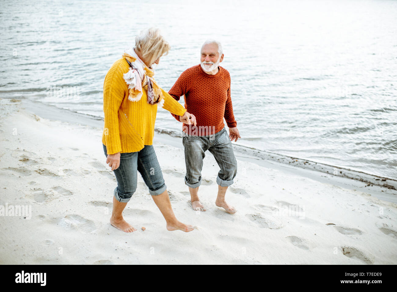 Lovely senior couple dressed in colorful sweaters walking on the sandy ...