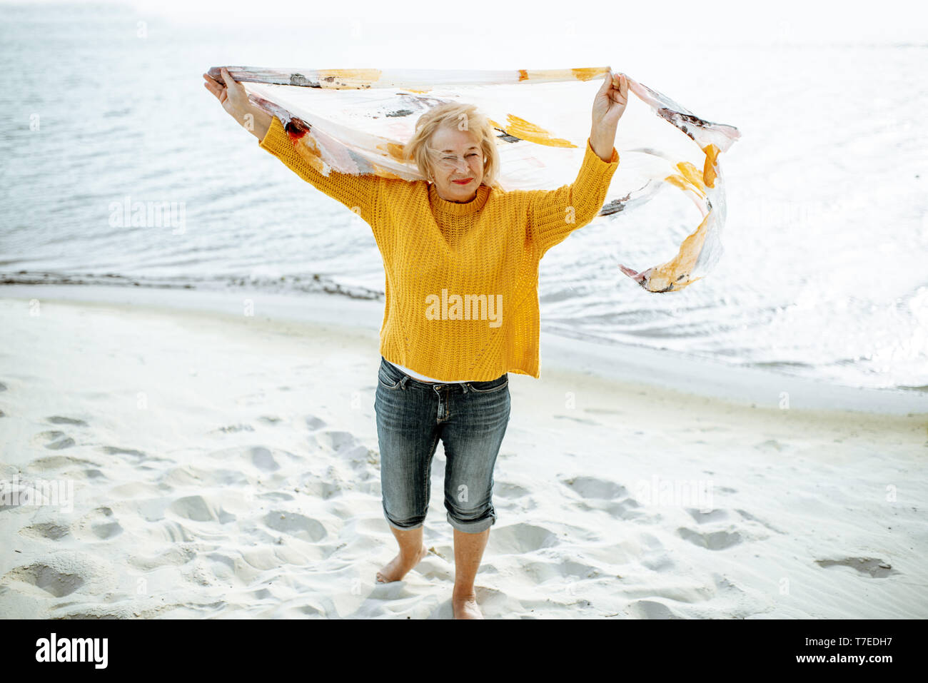Senior woman in bright sweater enjoying sea breeze, holding scarf above ...