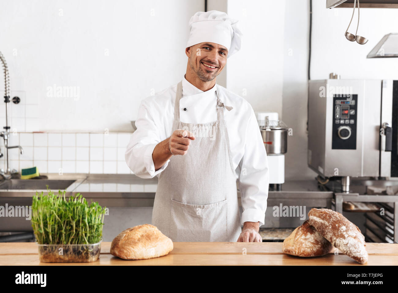 Image of positive man baker in white uniform smiling while standing at ...