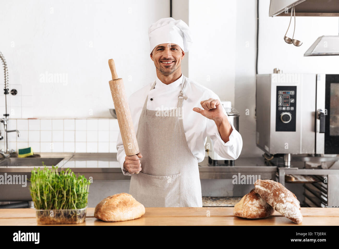 Image of joyful man baker in white uniform smiling while standing at ...