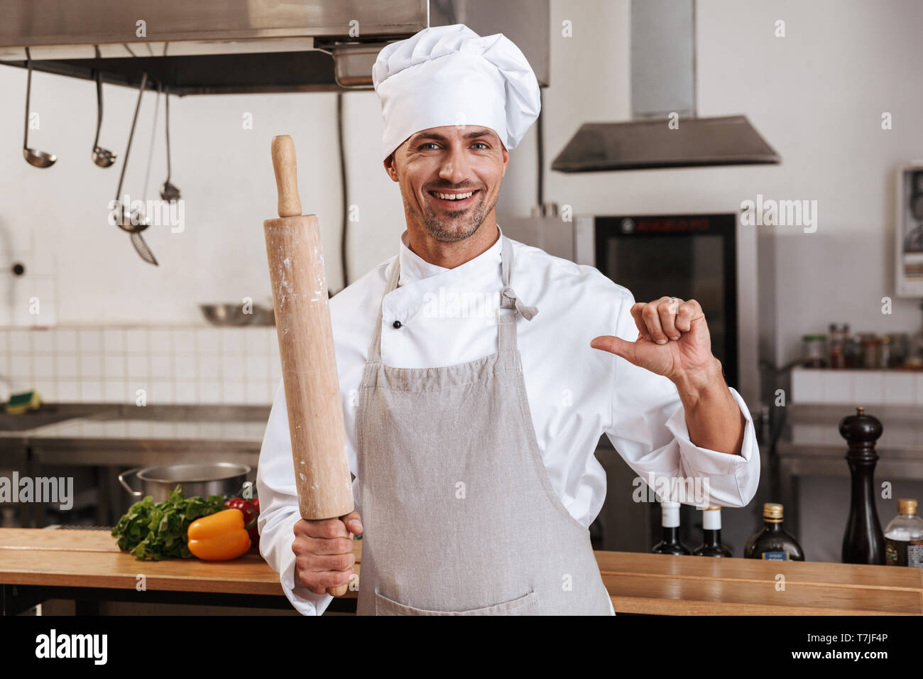 Photo of pleased male chief in white uniform holding rolling pin while ...
