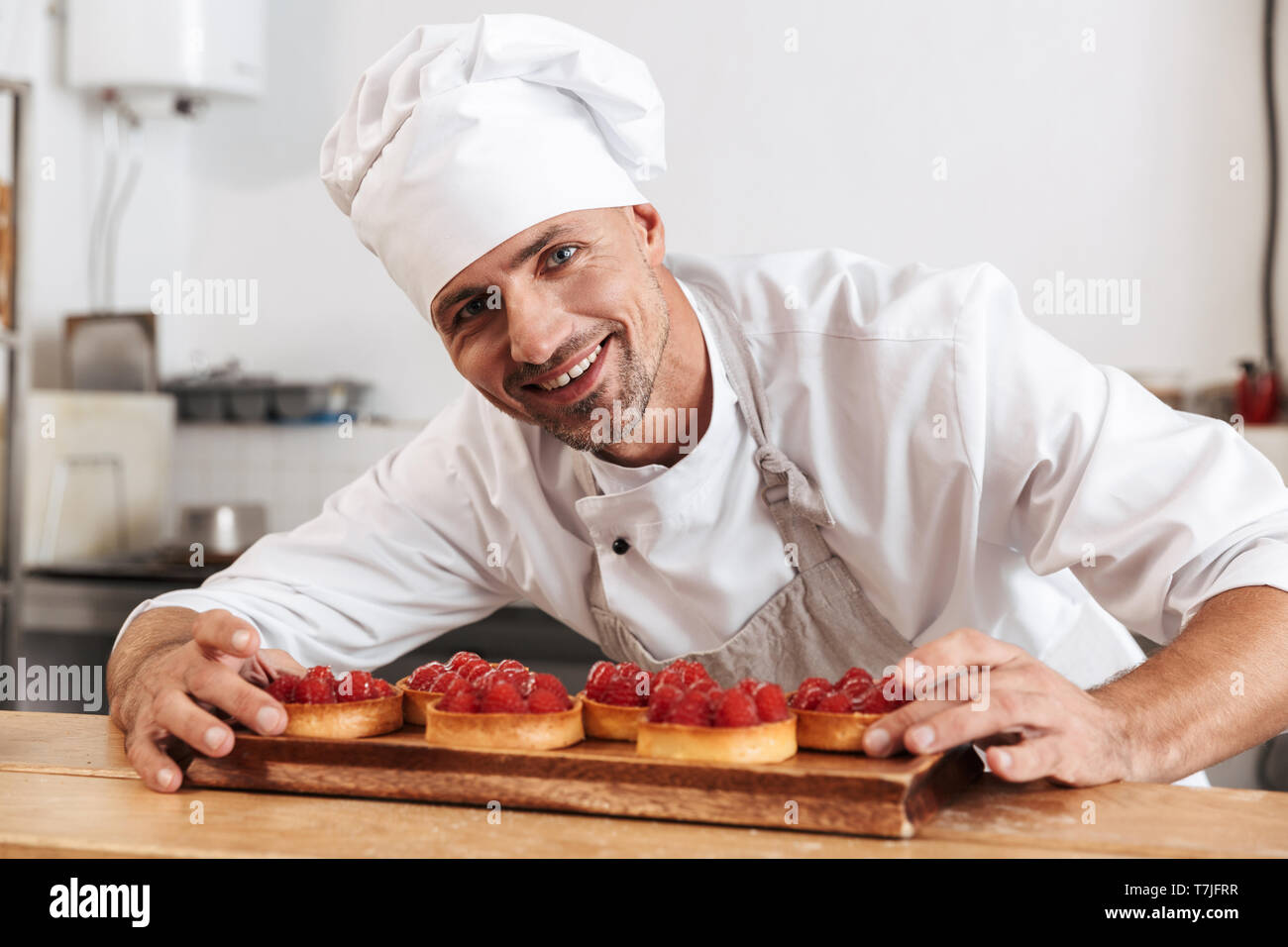 Photo of pleased male chief in white uniform holding plate with cakes ...