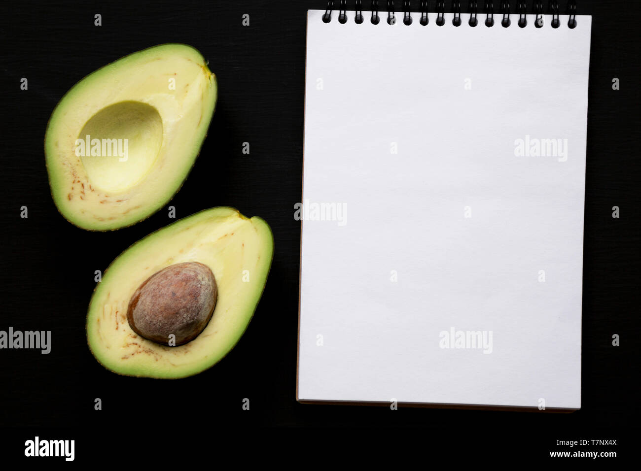 Halved avocado and blank notepad over black background, top view. From ...