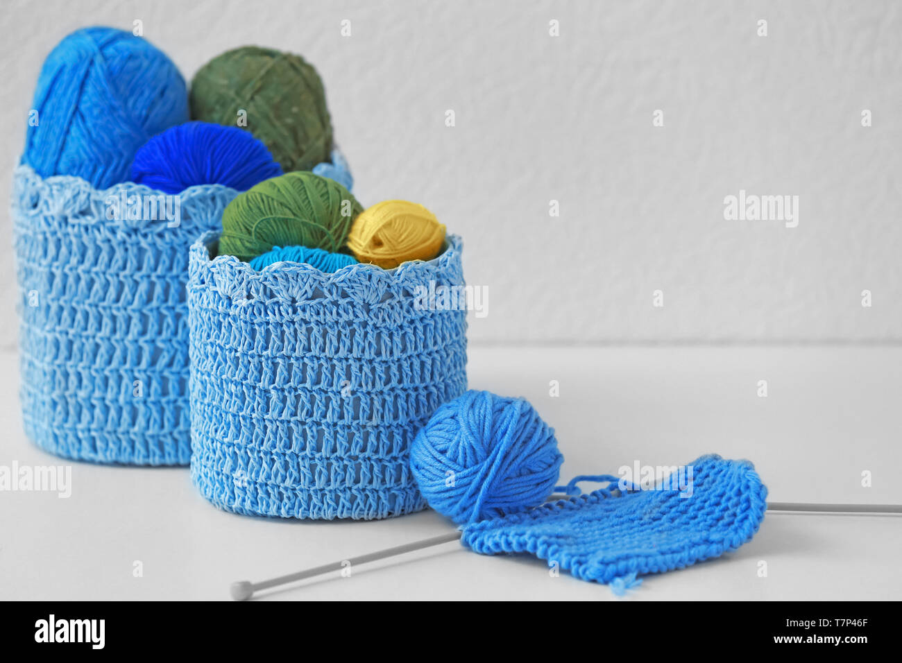 Basket with knitting yarn and unfinished clothes on table against light ...
