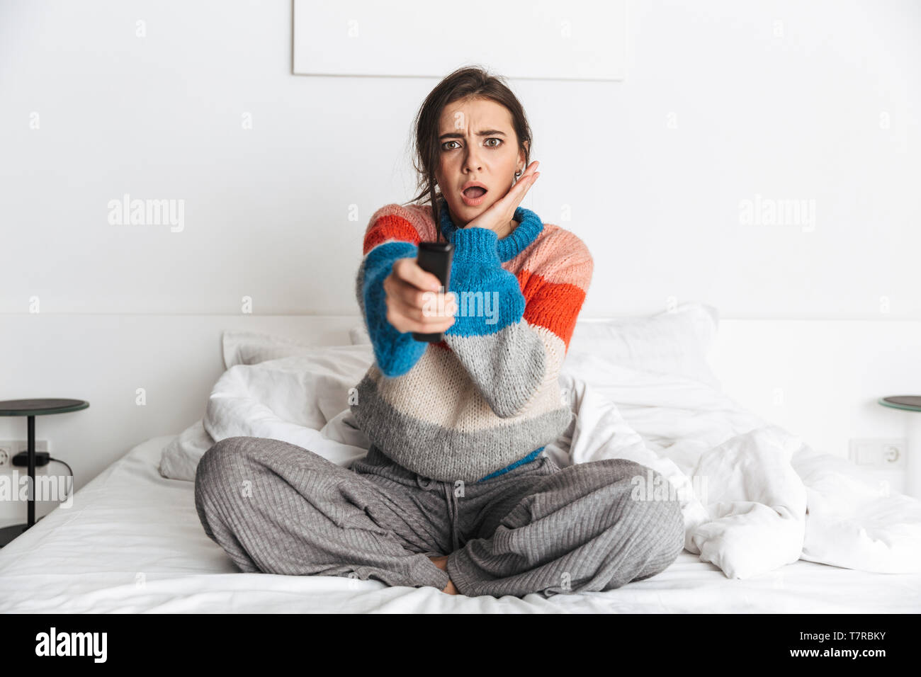 Portrait of surprised woman 20s holding remote control while sitting in ...